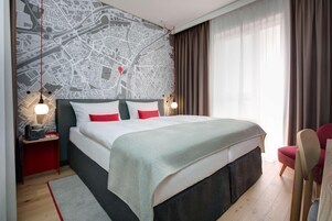 Quarto de hotel moderno com uma cama grande, uma cadeira vermelha, um mapa na parede e uma mesa de cabeceira com telefone.