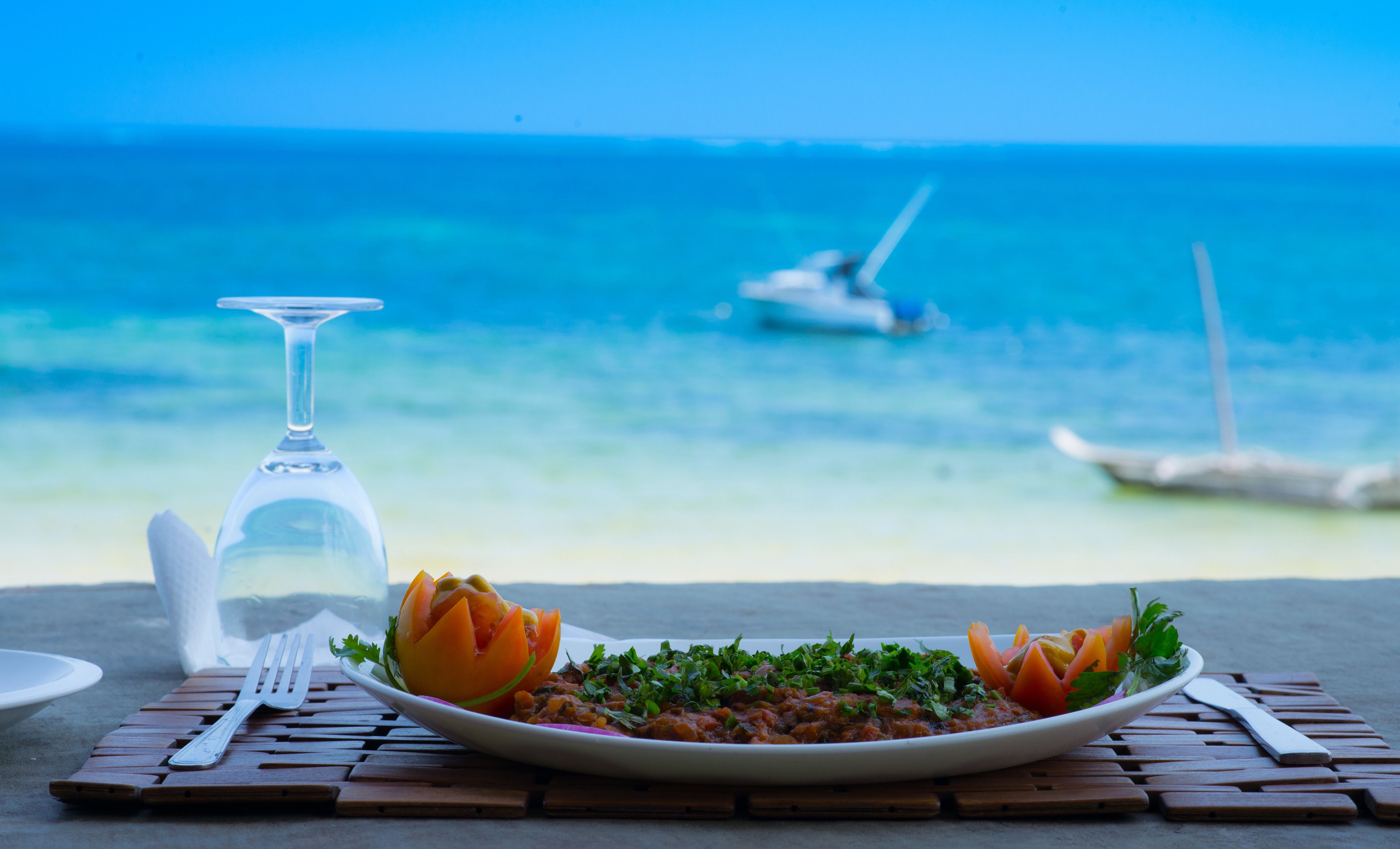 Uma mesa com prato de comida, taça de vinho e talheres, com vista para o mar e barcos.