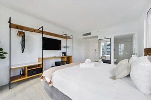 Quarto de hotel moderno com uma cama grande, uma TV montada numa estante de madeira, um banheiro visível por uma porta aberta e uma planta no canto.