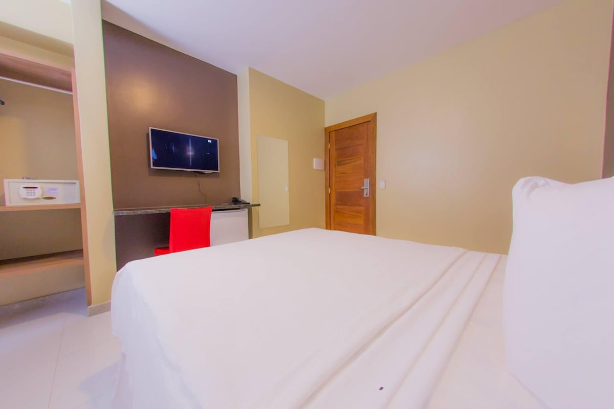 Quarto de hotel com cama, escrivaninha, televisão e porta.