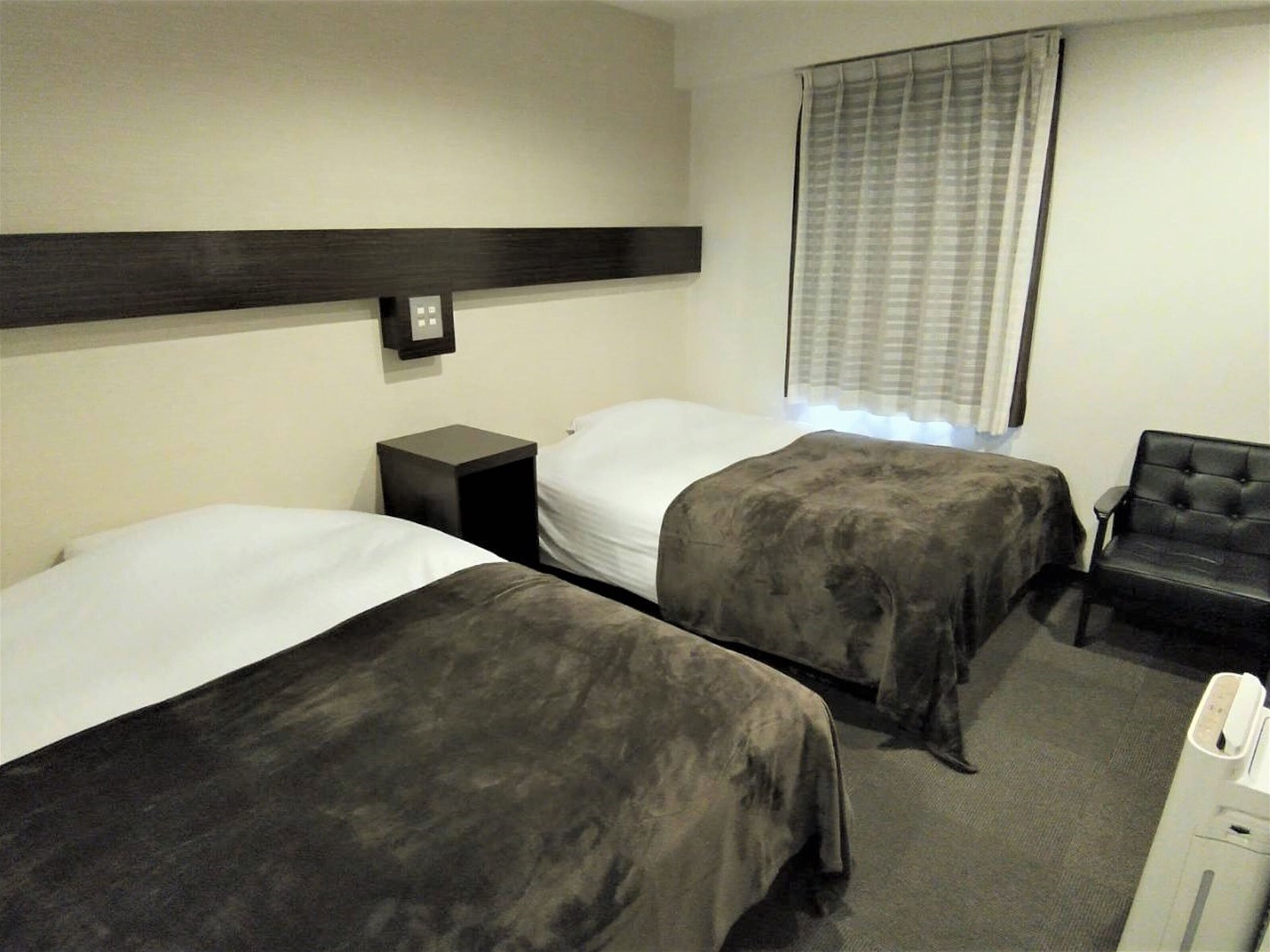 Quarto de hotel com duas camas, cabeceira escura, mesa de cabeceira pequena, uma cadeira e janela com persianas.