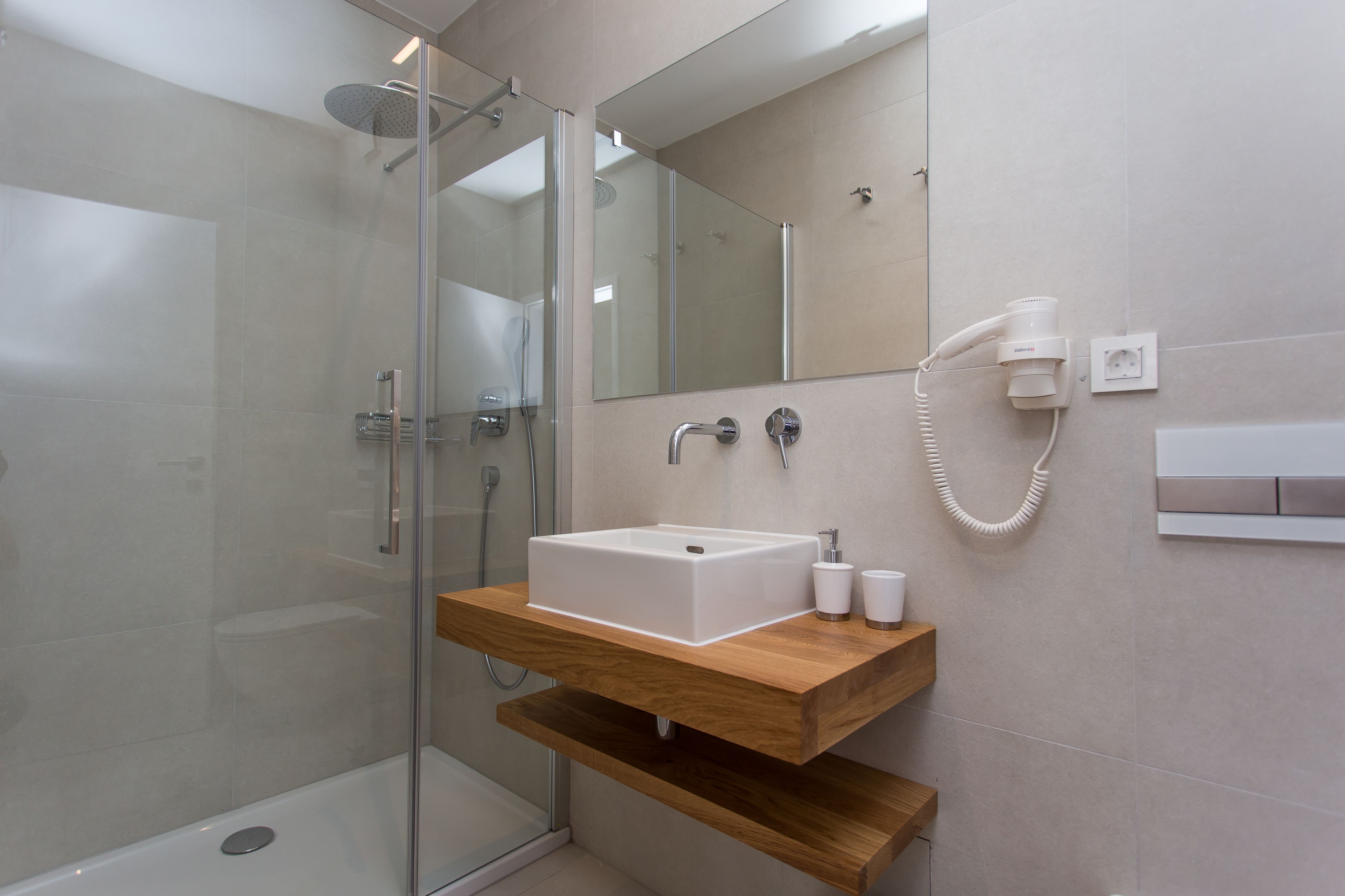 Uma casa de banho moderna com chuveiro em cabine de vidro, toucador de madeira com lavatório branco e secador de cabelo na parede.