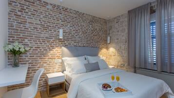 Quarto moderno com parede de tijolos, cama com roupa de cama branca, mesa de cabeceira, uma cadeira e um apoio para café da manhã com croissants e suco.