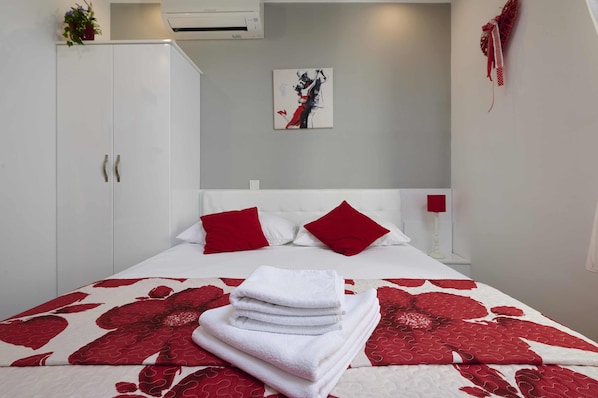 Cama bem arrumada com colcha floral vermelha e branca, pilha de toalhas brancas e dois travesseiros vermelhos.