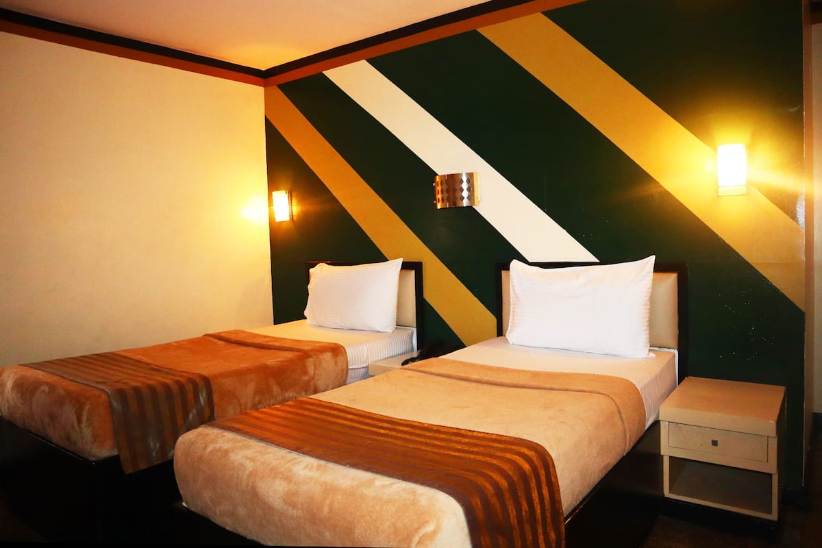 Quarto de hotel com duas camas, cada uma com travesseiro e cobertor. Há uma mesinha de cabeceira com uma luminária. As paredes têm um padrão listrado diagonal.