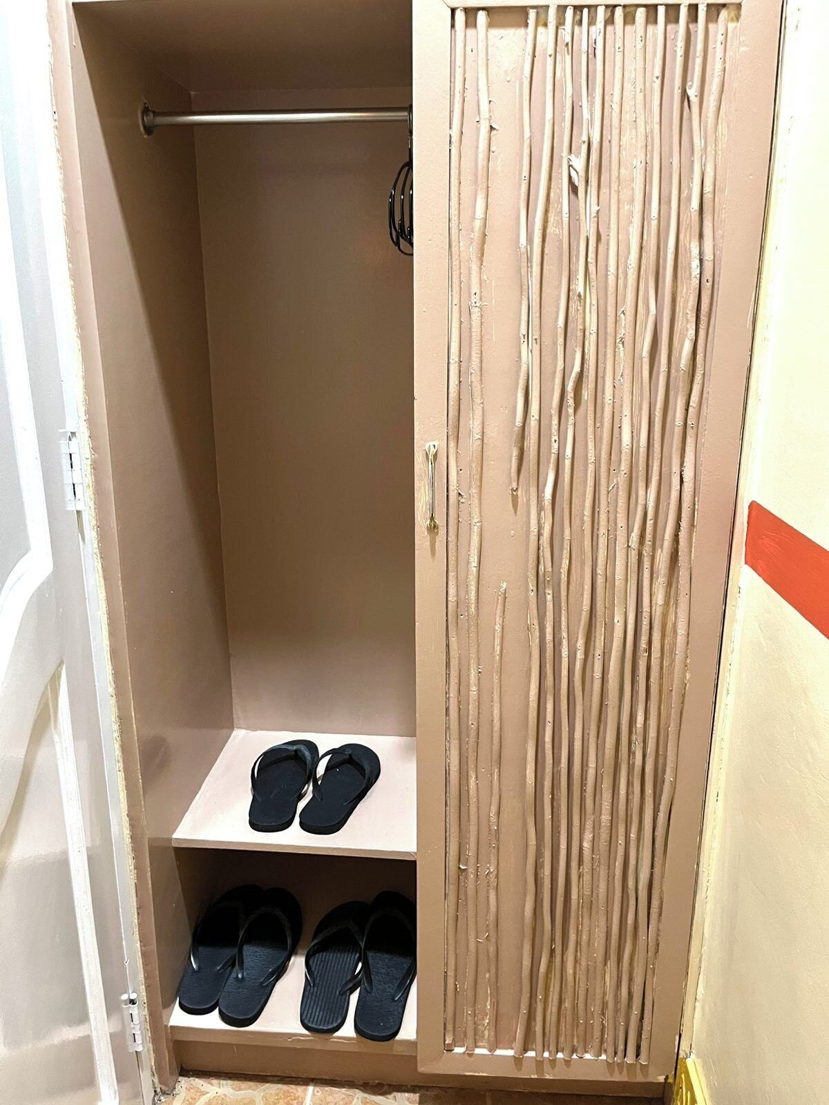 Casa de banho pequena e fechada, com uma prateleira que exibe chinelos pretos, um porta-toalhas e uma porta com tinta descascada.