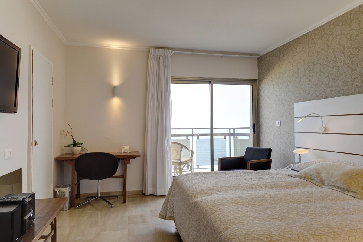 Quarto de hotel com cama, escrivaninha, cadeira, televisão e vista para o mar.