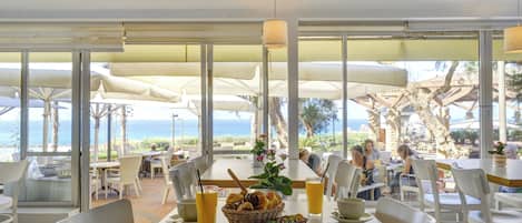 Um restaurante com mesas ao ar livre, grandes janelas e vista para o mar.