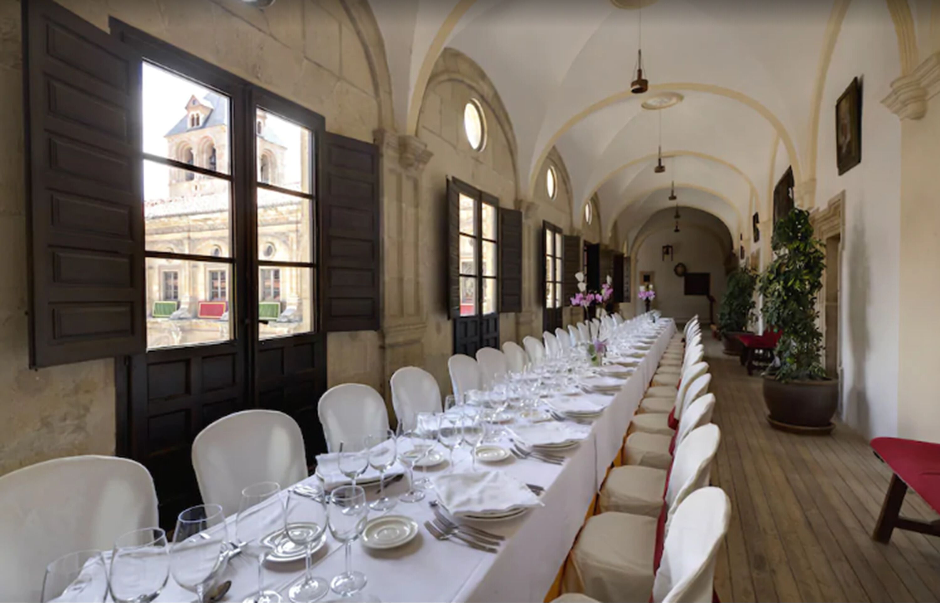 Uma longa mesa de jantar posta com cadeiras brancas, amplas janelas com persianas e vista para um edifício.