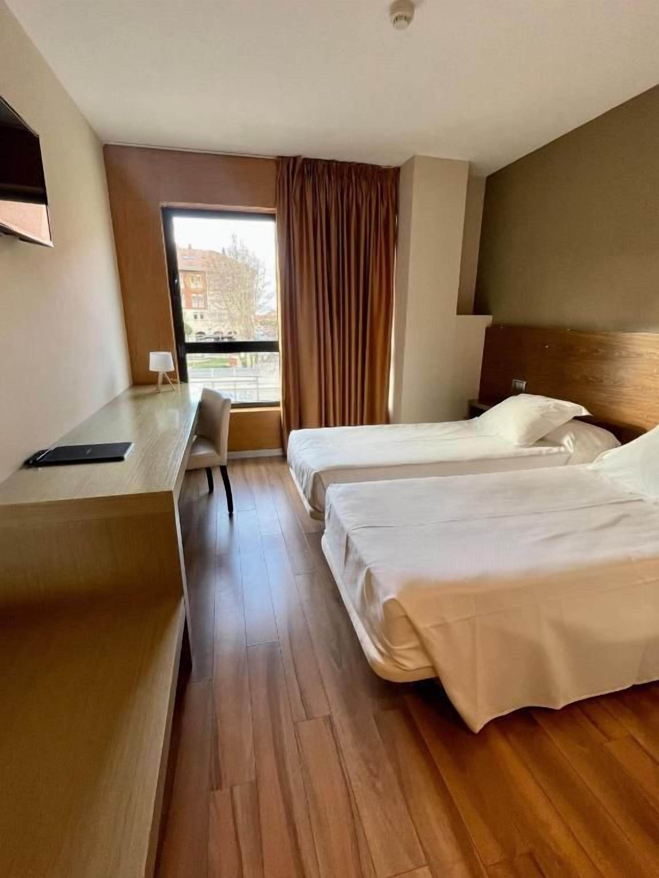 Quarto de hotel com duas camas, uma escrivaninha e janela com cortinas.