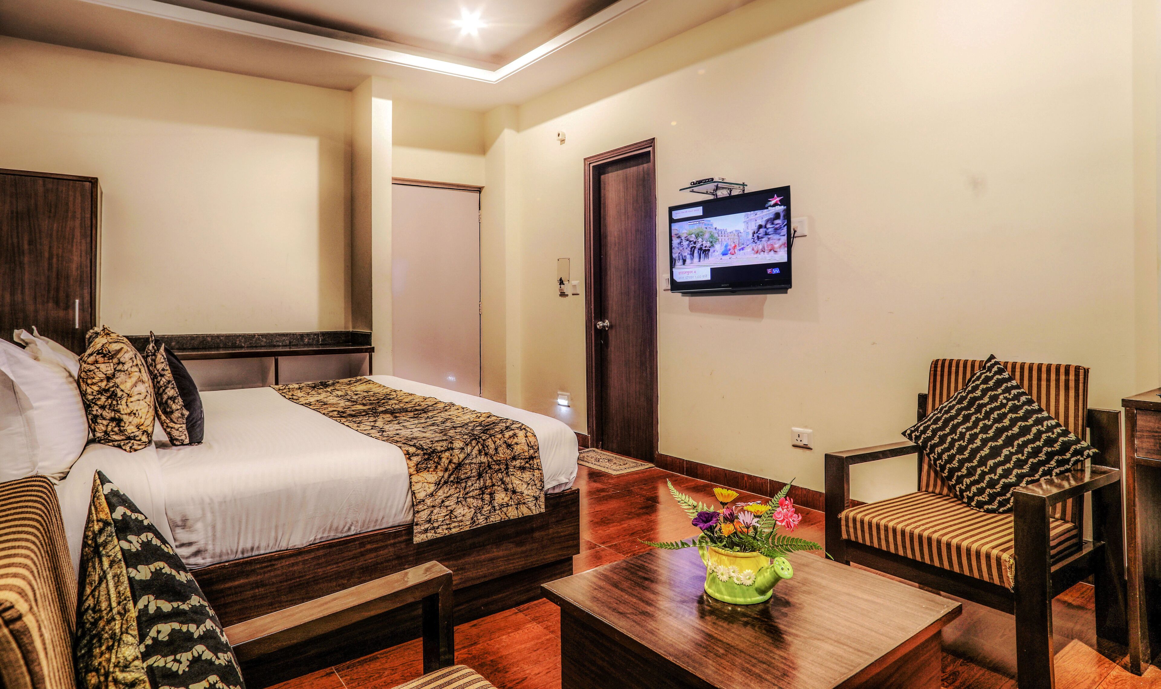 Quarto de hotel com cama, sofá, poltrona, televisão e uma pequena mesa com um vaso de flores.