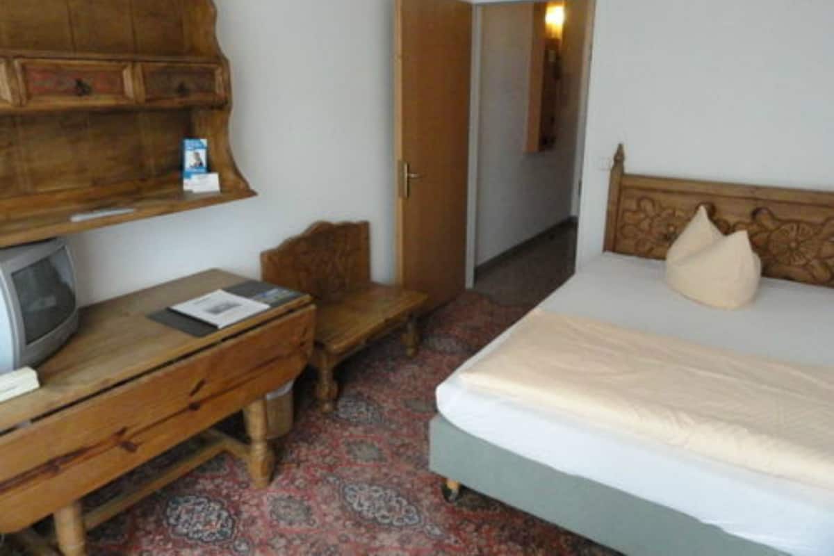 Um quarto com cama, escrivaninha de madeira com computador, uma cadeira e guarda-roupa.
