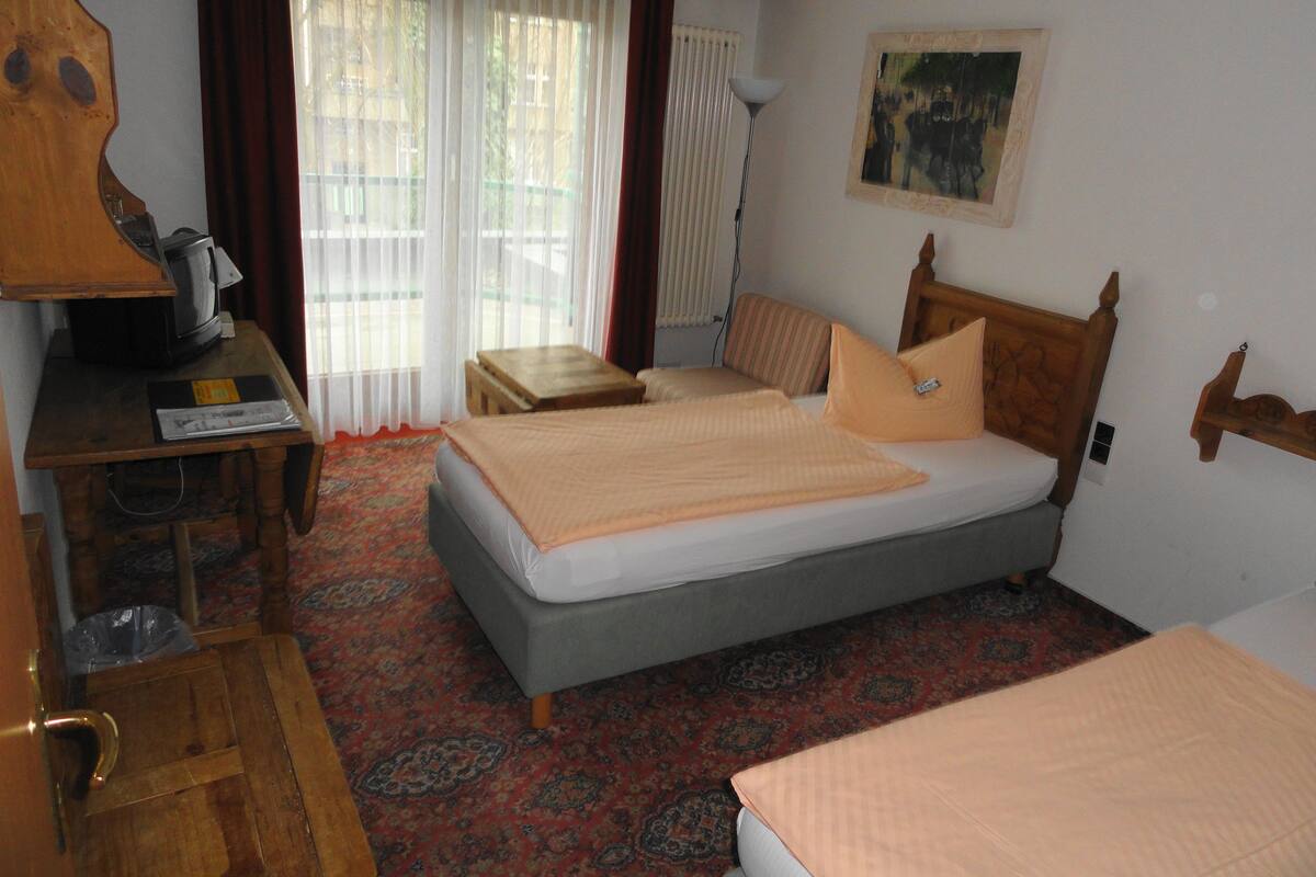Quarto de hotel com cama, escrivaninha com computador, cadeira e janela com cortinas.