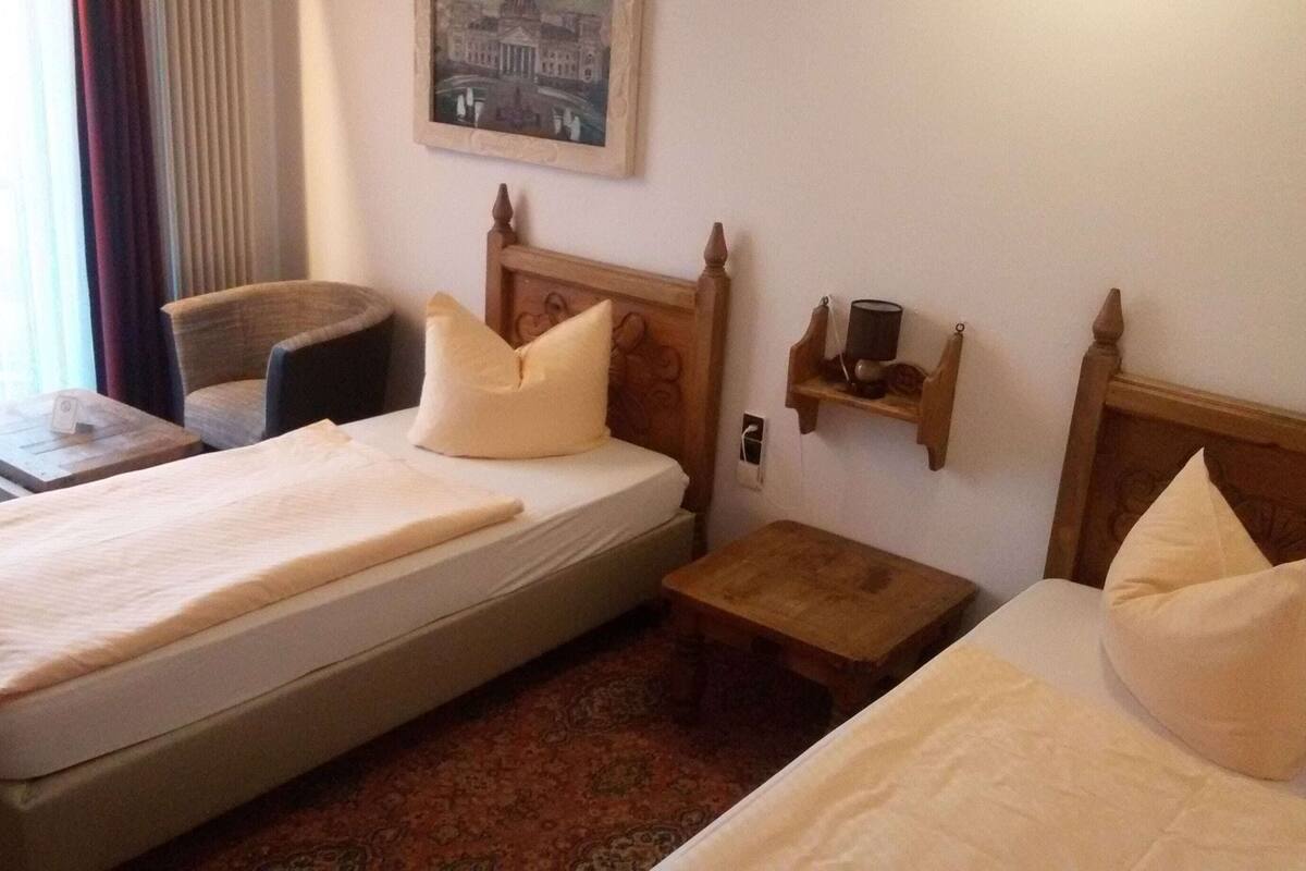Quarto de hotel com cama, uma cadeira, uma mesinha, mesa de cabeceira e prateleira na parede.