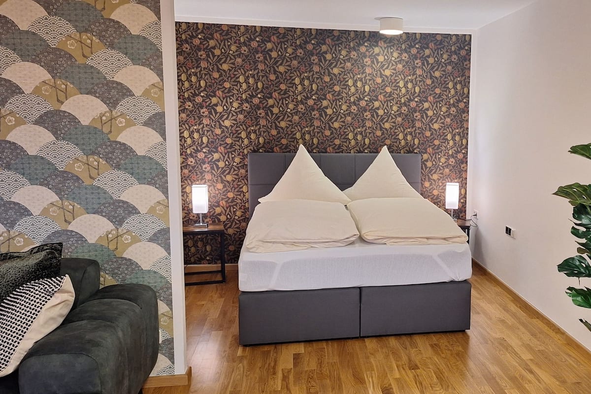 Quarto com cama, mesa de cabeceira, luminária, planta e porta de madeira.