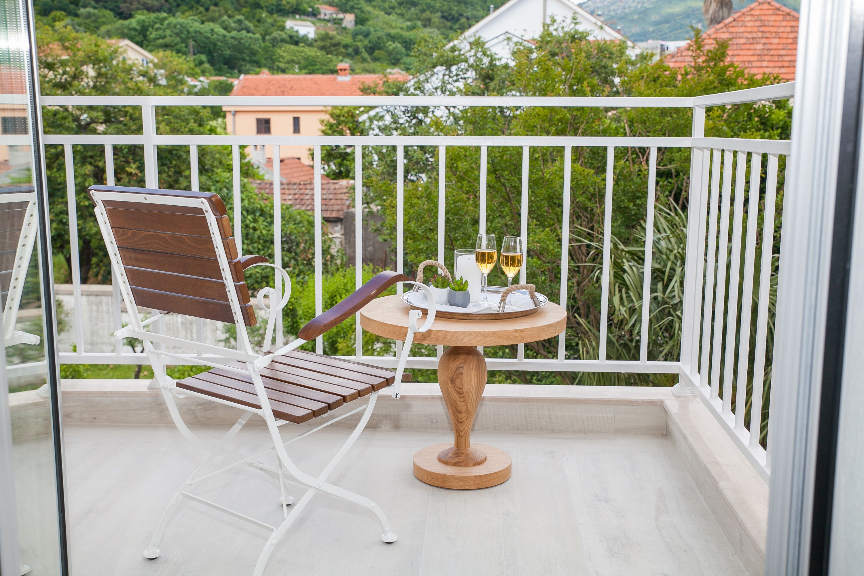 Varanda com corrimão branco, uma cadeira de madeira, uma mesa redonda pequena com uma taça de vinho e uma garrafa, e vista para o verde e edifícios ao fundo.
