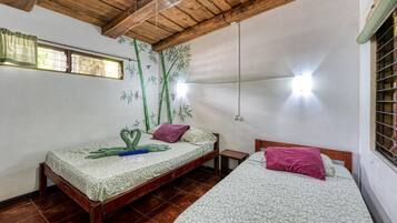 Quarto com duas camas, vigas de madeira no teto, arte de parede em bambu e janela com grades.