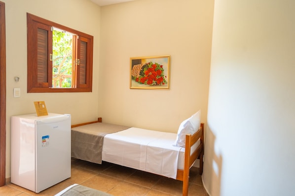 Quarto pequeno e compacto com uma cama de solteiro, um refrigerador, uma janela com persianas de madeira e um quadro com rosas vermelhas na parede.