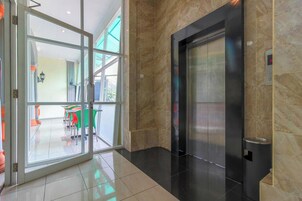 Um elevador moderno, entrada com porta de vidro e vista para uma área externa com mesas e cadeiras.