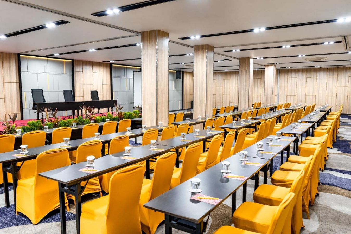Sala de conferências com fileiras de mesas e cadeiras, um palco com piano e uma janela grande.