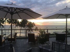 Terraço com mesas ao ar livre, palmeiras e vista para o oceano ao pôr do sol.