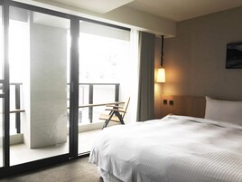 Quarto de hotel com uma cama, uma cadeira, varanda e luminária fixa na parede.