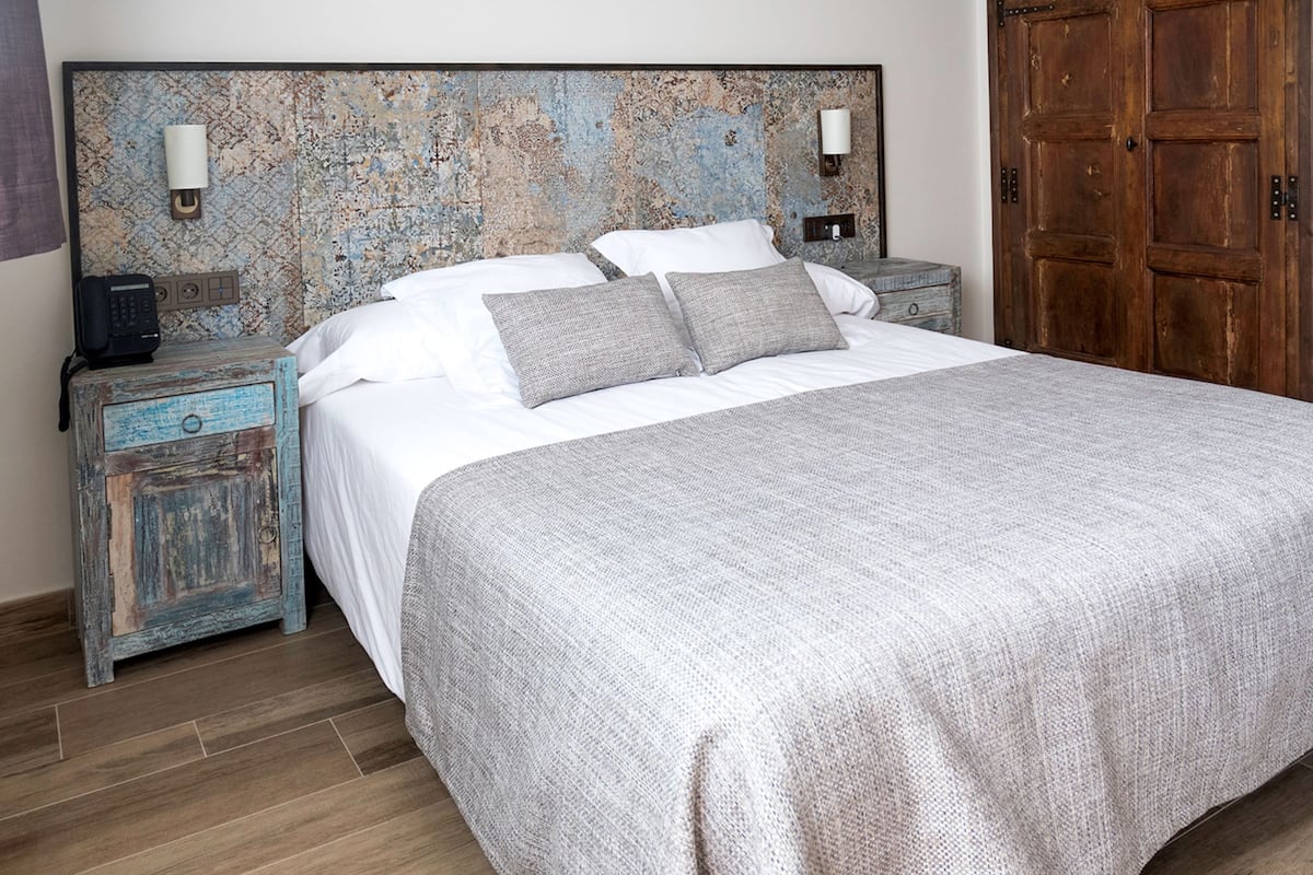 Cama com cabeceira texturizada, roupa de cama branca e duas almofadas. Apresenta uma mesa de cabeceira com uma luminária e um telefone. Guarda-roupa de madeira visível à direita.
