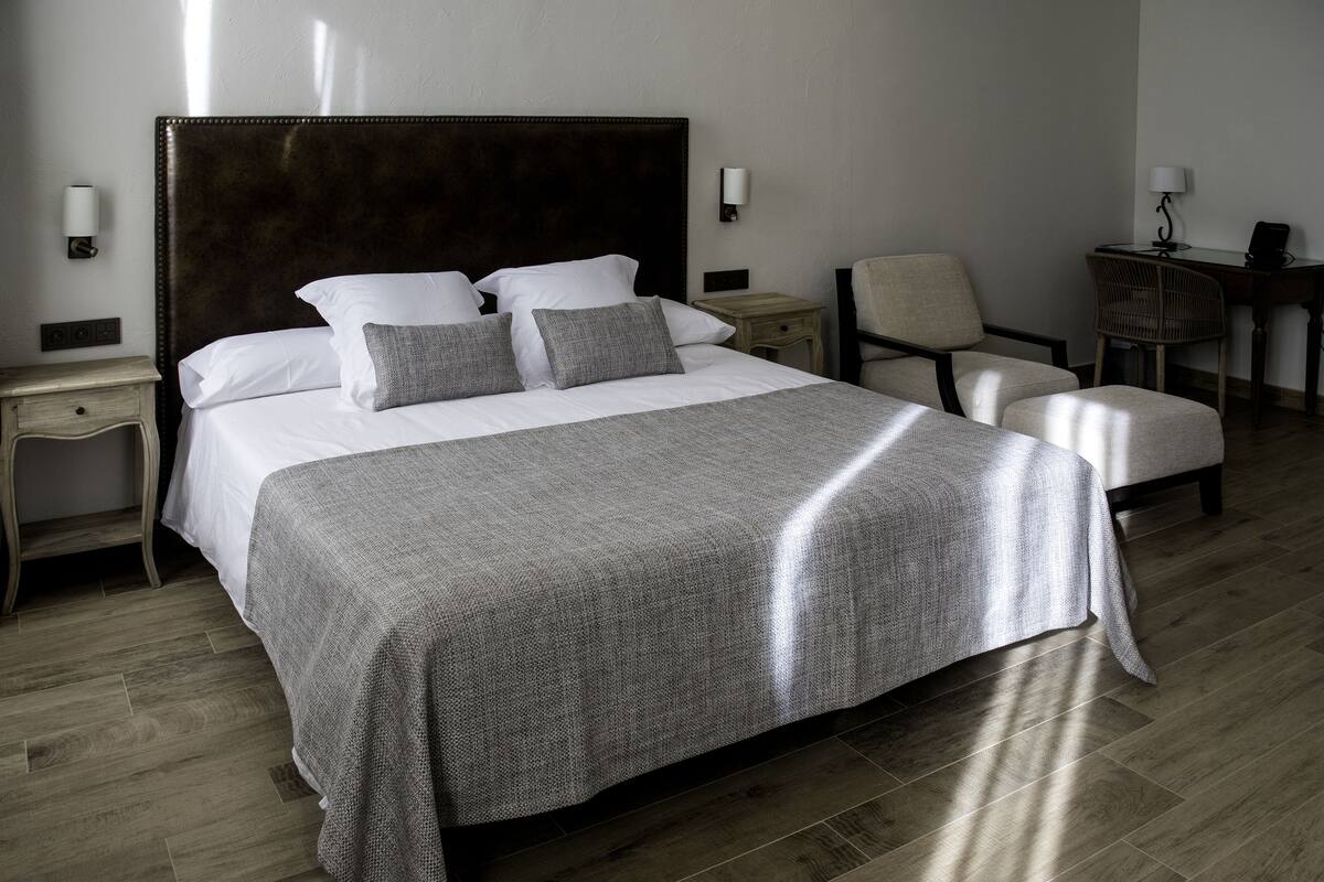 Uma cama bem arrumada com um cobertor cinza, uma cabeceira de madeira e duas mesas de cabeceira com abajures.