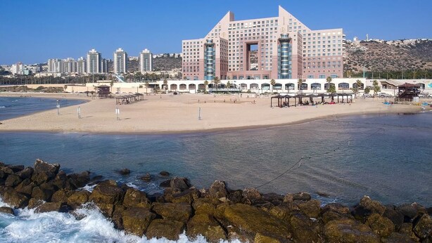 Um hotel grande com um pátio central, uma praia ao fundo e uma linha costeira rochosa em primeiro plano.