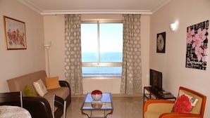 Uma sala de estar com sofá, poltrona, mesa de centro e televisão. Há uma janela com cortinas e vista para o mar.