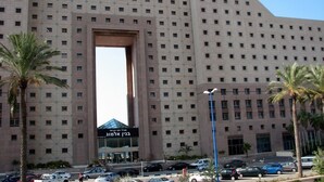 Um edifício grande com uma entrada em arco marcante, vários andares e um estacionamento com carros e palmeiras.