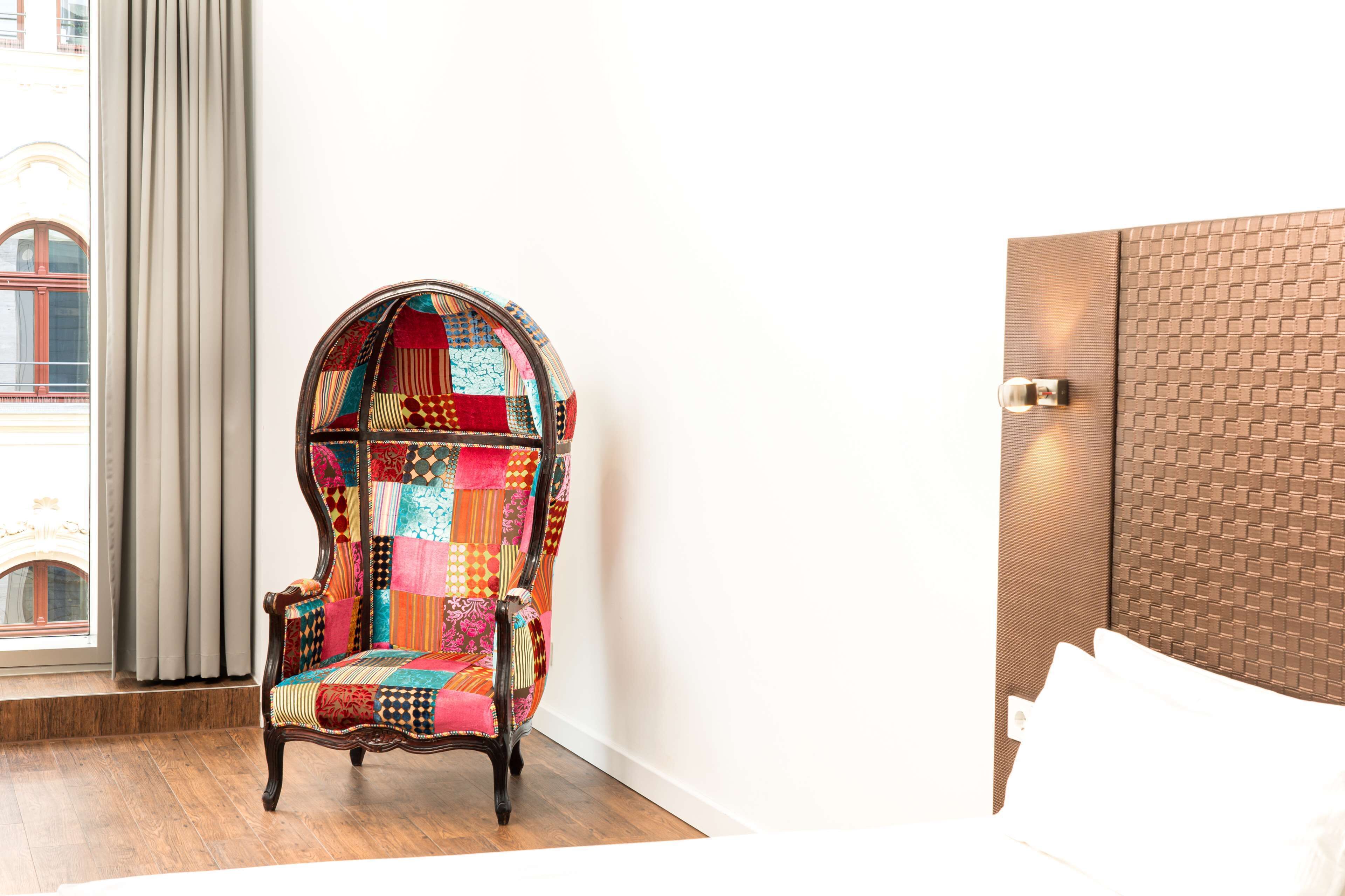 Um quarto com piso de madeira, uma poltrona colorida, uma cama com travesseiros brancos e uma cabeceira com padrão de tijolos.
