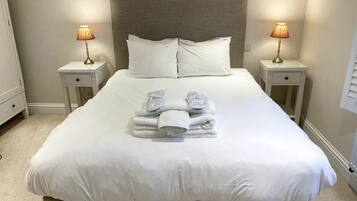 Cama bem arrumada com lençóis brancos, duas mesas de cabeceira com luminárias e uma pilha de toalhas dobradas na cama.
