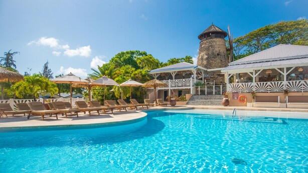 Uma área de piscina com espreguiçadeiras e guarda-sóis, uma torre de pedra e um edifício com telhado de palha.