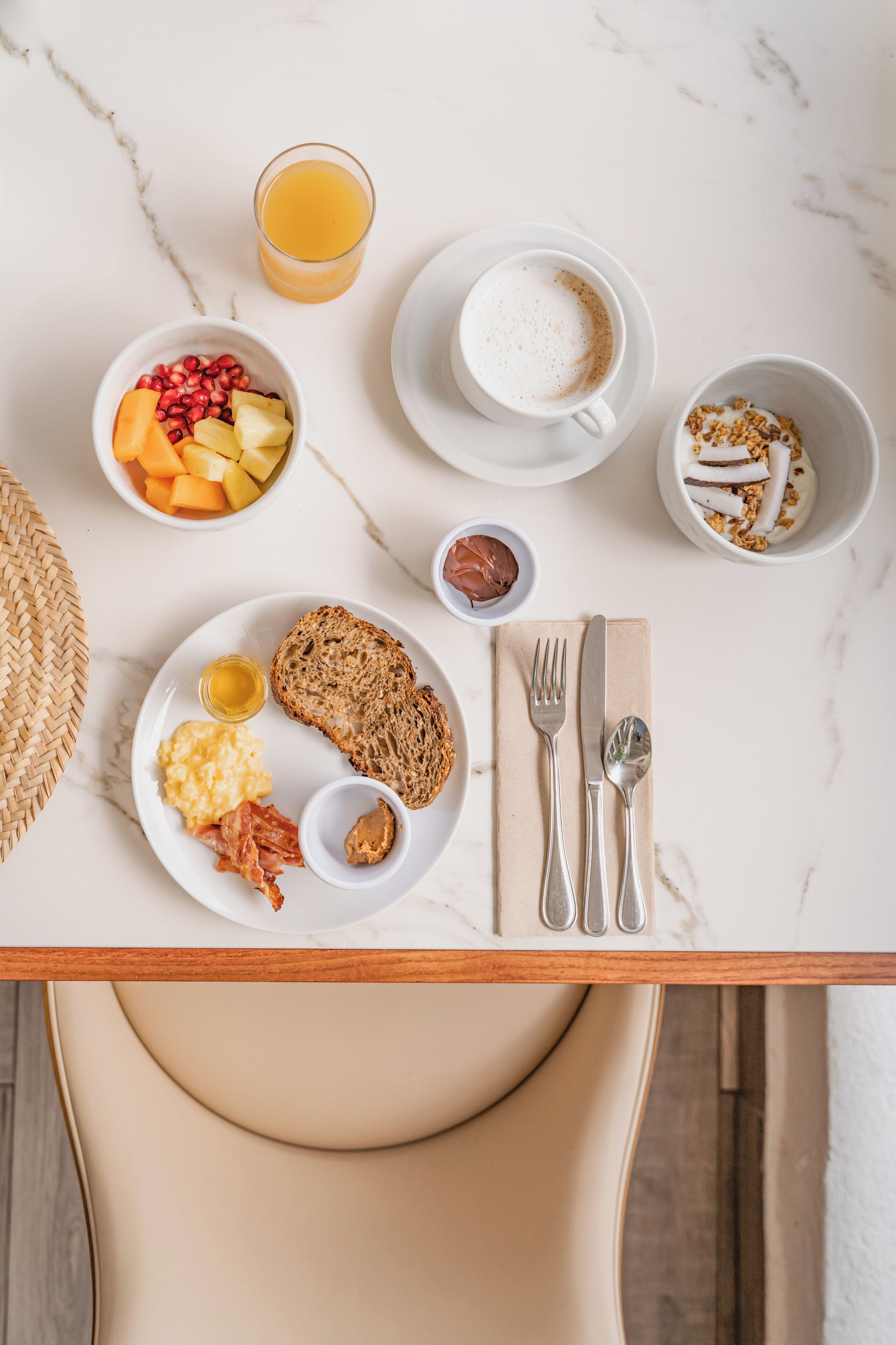 Mesa de pequeno-almoço com diversos alimentos, um copo de sumo de laranja e utensílios de mesa.