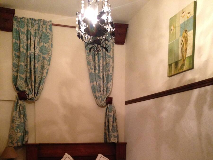 Quarto com cama, cortinas, mesa de cabeceira, abajur, cadeira e um quadro na parede.