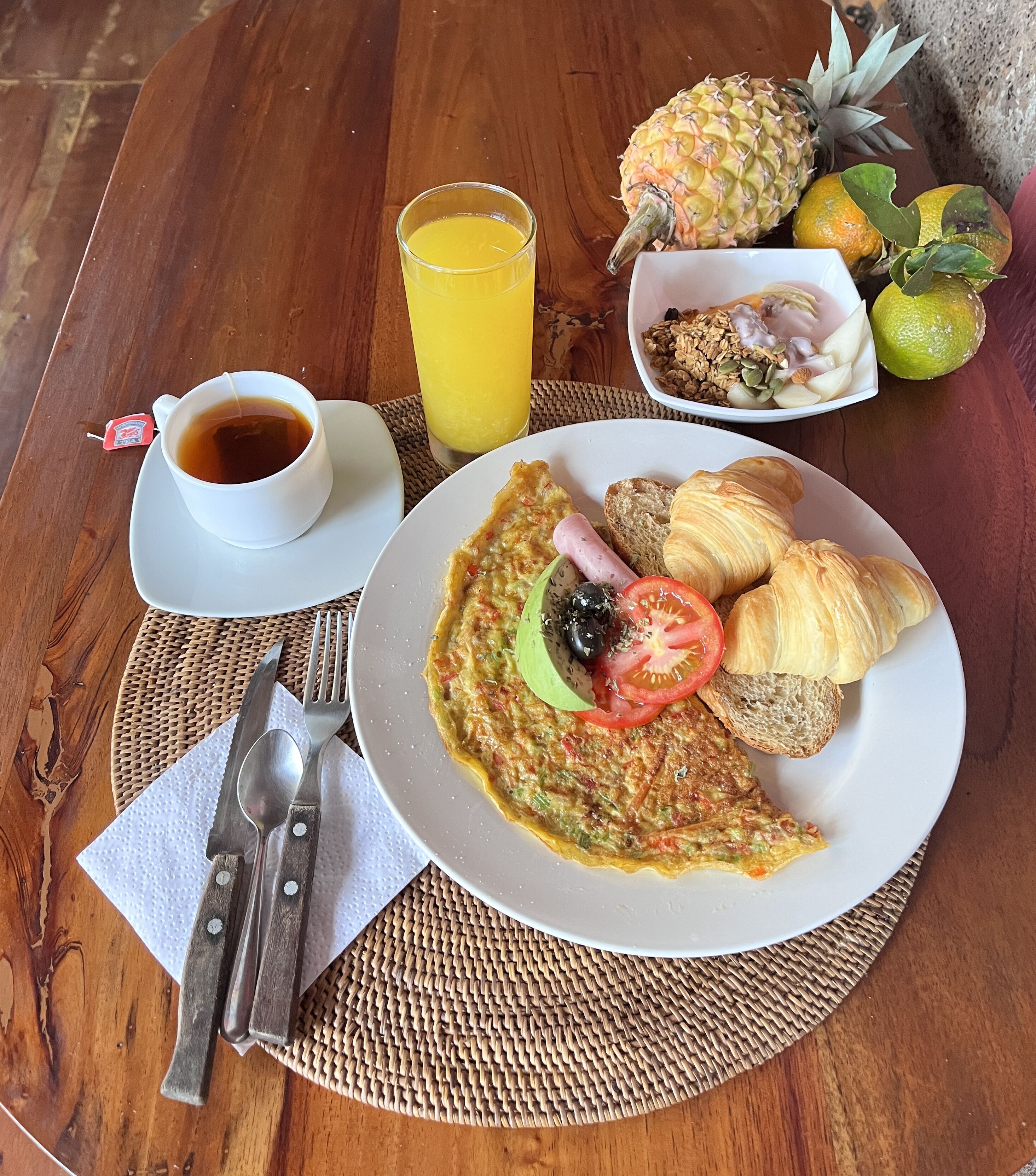 Mesa posta para o café da manhã, com prato de comida, copo de suco de laranja, xícara de café e uma abacaxi sobre a mesa.