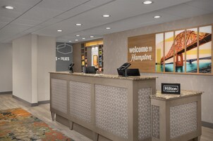 Uma receção de hotel com um letreiro 'Welcome to Hampton', um letreiro 'Hilton Honors' e um letreiro 'Treats'.