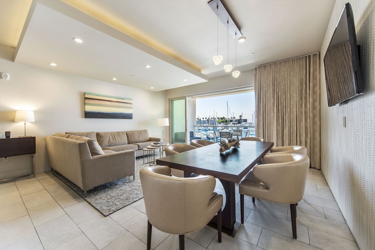 Uma sala de estar moderna com sofá, mesa de jantar e cadeiras, TV fixada na parede e vista para a marina através de grandes janelas.