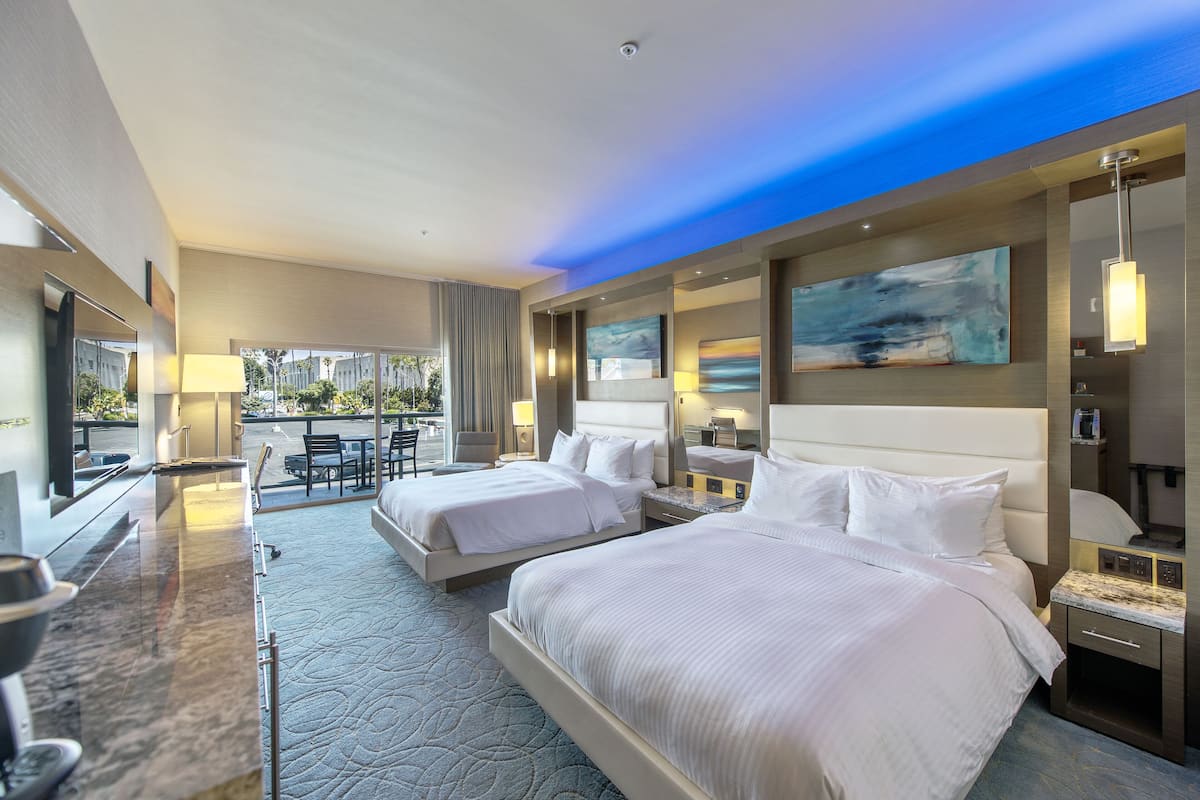 Quarto de hotel com duas camas, um espaço de estar com mesa e cadeiras, e obras de arte nas paredes.