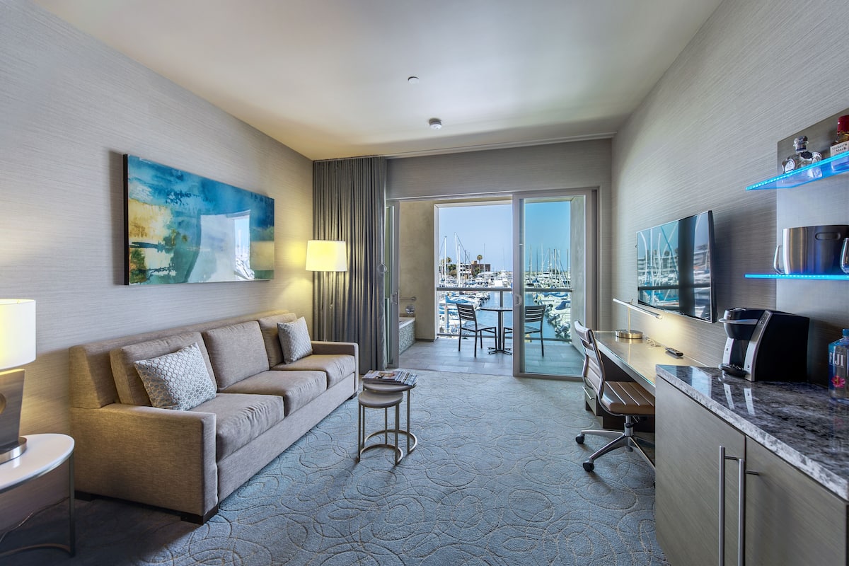 Quarto de hotel com sofá, uma pequena mesa, televisão, máquina de café e vista para a marina.