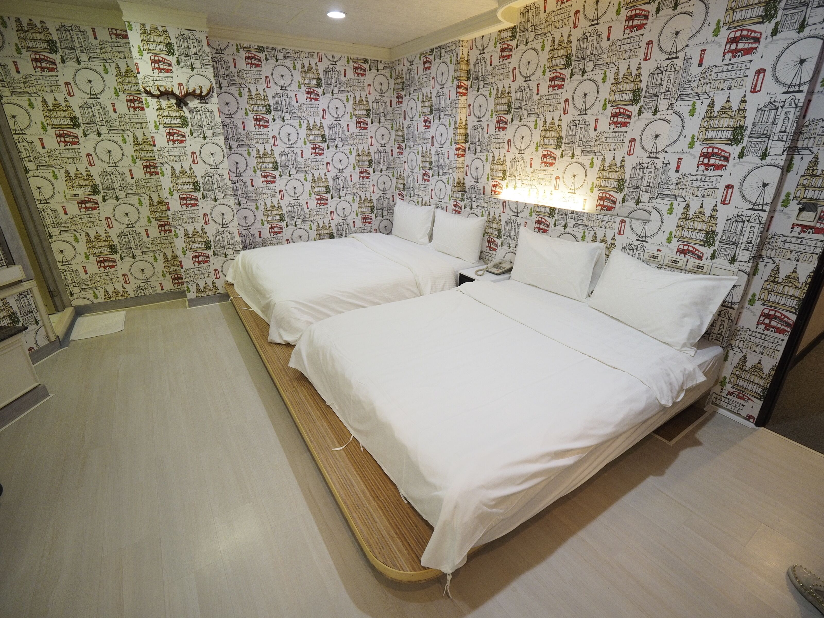 Quarto de hotel com duas camas, piso de madeira e papel de parede estampado com paisagens urbanas.