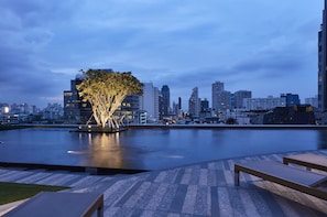 Paisagem urbana à beira-mar com edifícios modernos, uma árvore iluminada por luzes e um corpo d'água calmo.