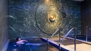 Área de spa com parede de mármore, luminária circular e uma pessoa relaxando em uma banheira de hidromassagem.