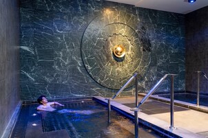 Área de spa com parede de mármore, luminária circular e uma pessoa relaxando em uma banheira de hidromassagem.