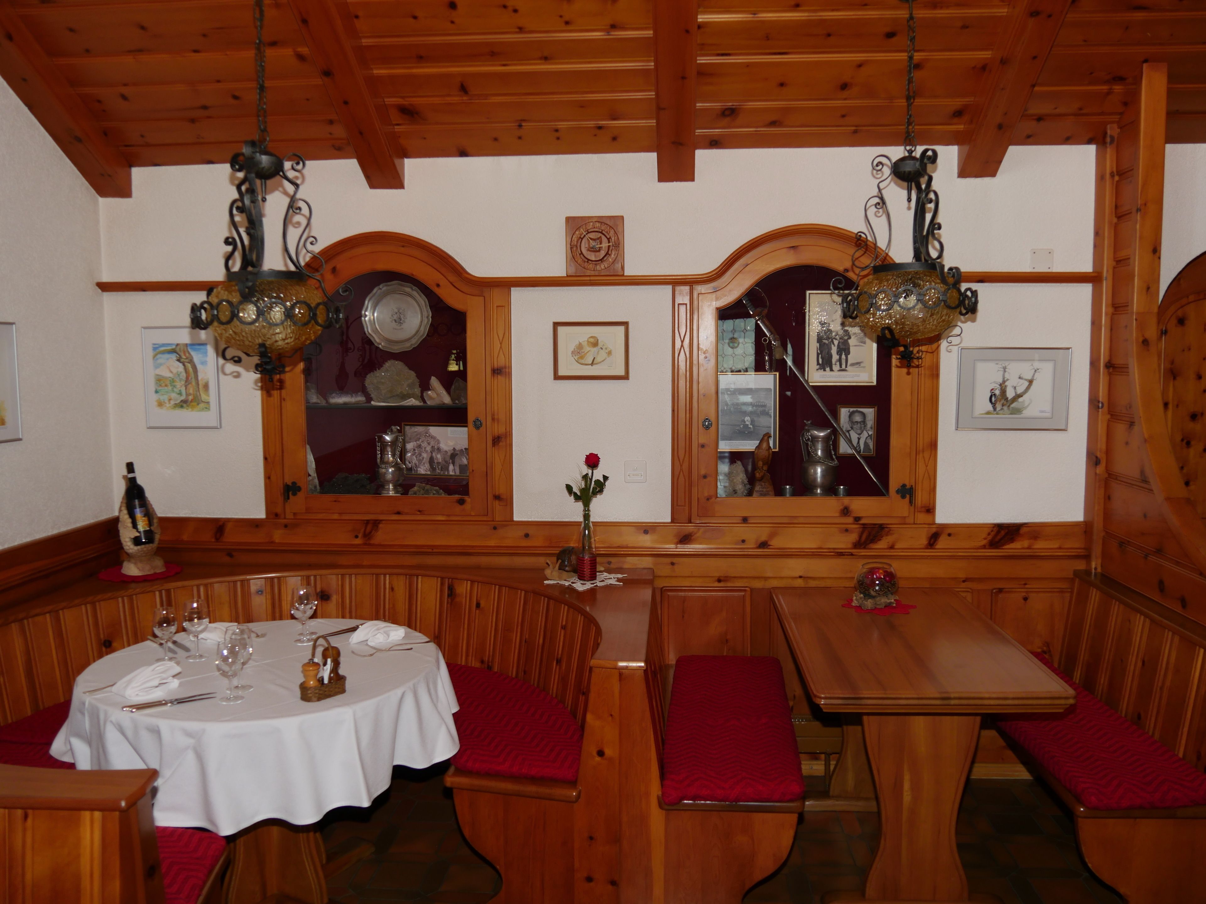 Uma área de refeições com mesa redonda posta para quatro pessoas, bancos com almofadas vermelhas e teto de madeira com vigas aparentes.