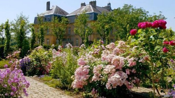 Um caminho no jardim leva a um edifício com telhado azul e várias chaminés.