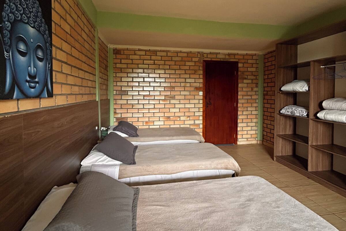Quarto com três camas, parede de tijolos, pintura de Buda e guarda-roupa de madeira.
