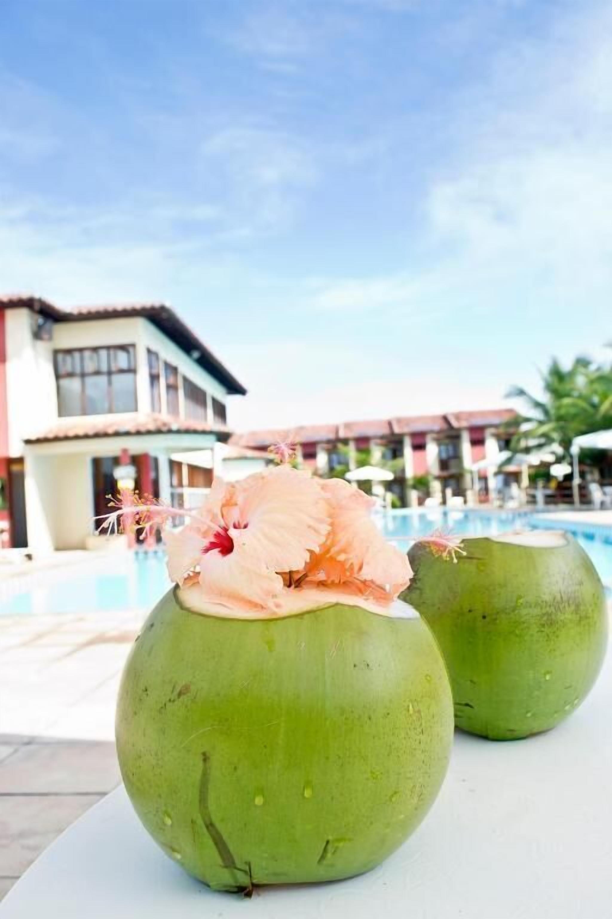 Área junto à piscina com dois cocos verdes sobre uma mesa branca, um edifício com varandas e céu claro.