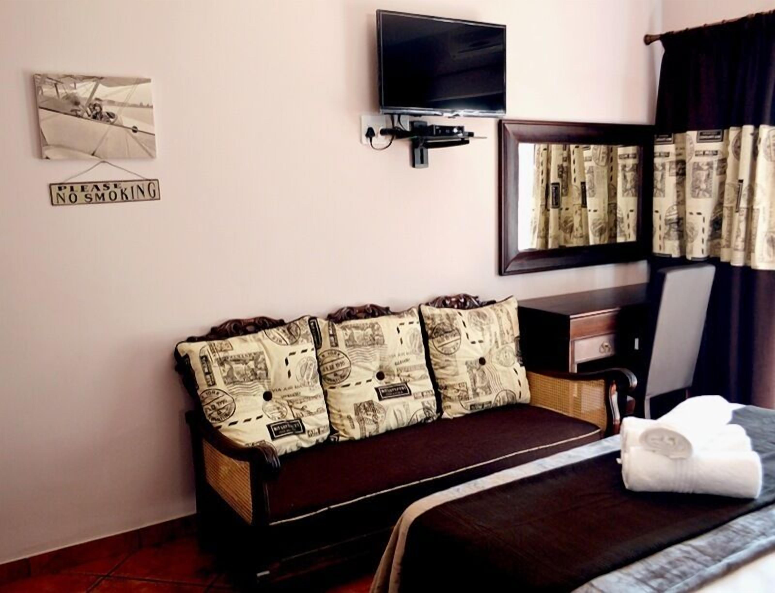 Quarto de hotel com cama, televisão fixada na parede, sofá com almofadas estampadas com cifrões e um aviso de 'NÃO FUMAR' na parede.